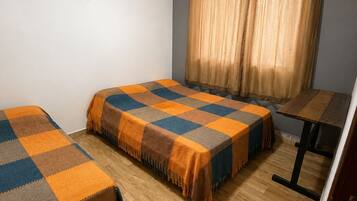 Quarto com duas camas, ventilador de teto, janela com cortinas e uma pequena mesa.