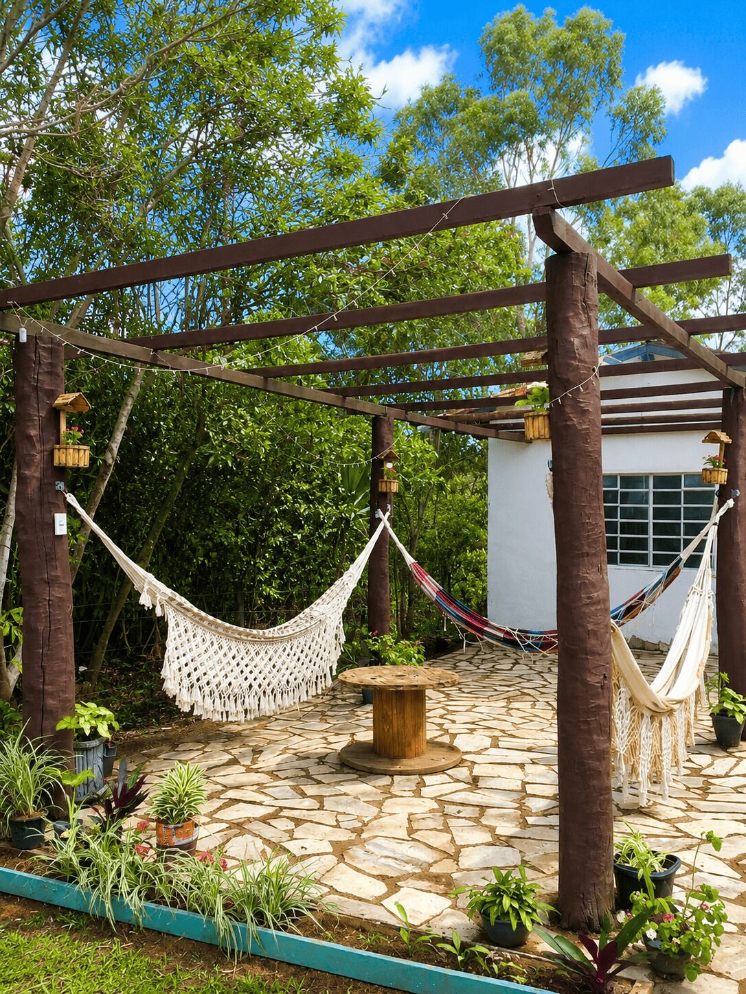 Uma rede sob uma pérgola de madeira, rodeada por vasos de plantas e um pátio de pedra.