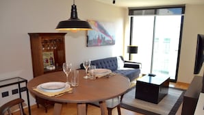Uma sala de jantar com mesa redonda posta para quatro pessoas, um sofá escuro, uma televisão e uma pintura com vista para a cidade.
