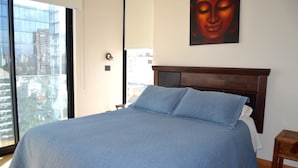Quarto com uma cama grande, cabeceira de madeira, janela com vista para prédios e uma estátua de Buda na parede.