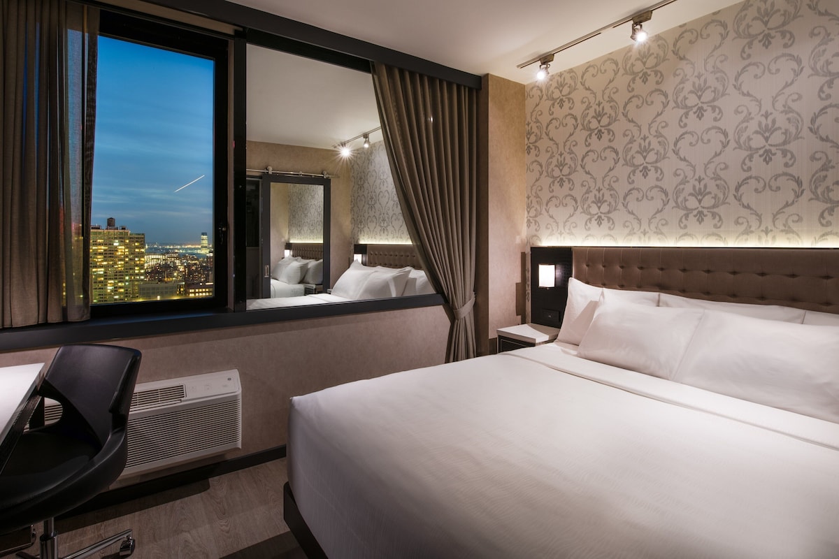 Quarto de hotel com uma cama grande, uma mesa, uma cadeira, um espelho e vista da cidade à noite.