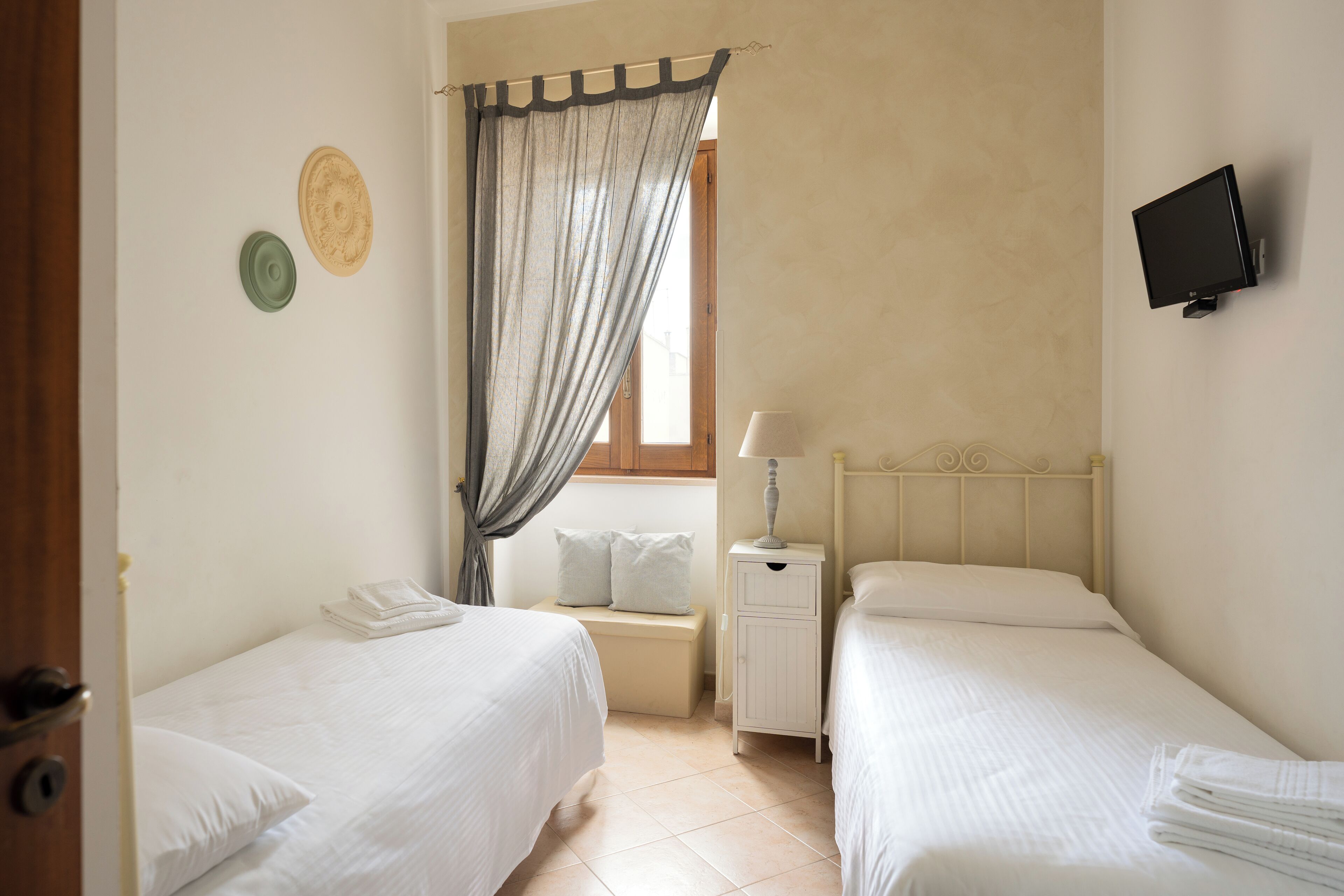 Quarto de hotel com duas camas, mesa de cabeceira, luminária, televisão e janela com cortinas.