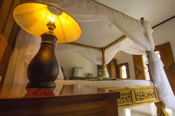 Uma cama com dossel, uma mesa de cabeceira com abajur e uma mesinha de cabeceira com rádio.