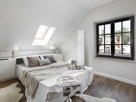 Quarto com teto inclinado, claraboia, cama com cabeceira branca, mesas de cabeceira com luminárias e vista para edifícios por uma janela grande.