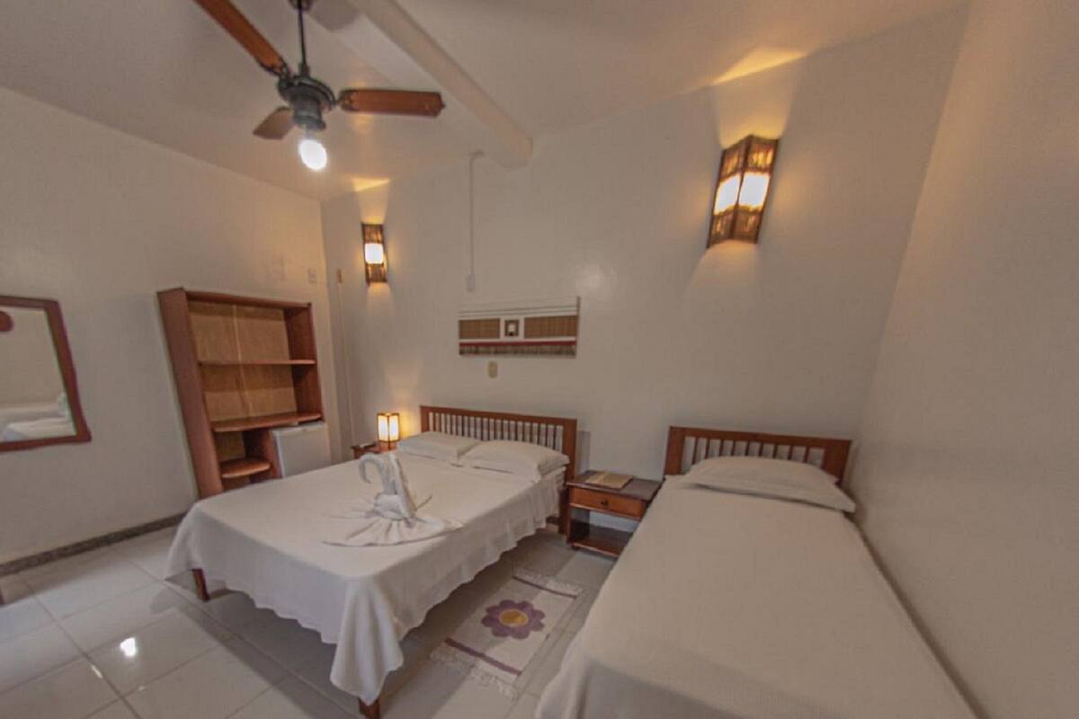 Quarto com duas camas, ventoinha no teto, mesa de cabeceira com luminária, guarda-roupa e espelho.