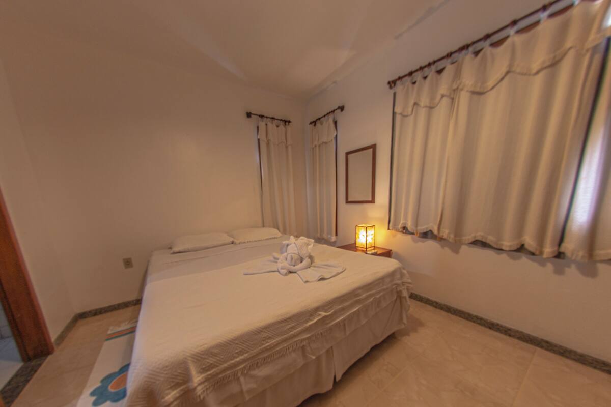 Quarto de hotel com uma cama grande, uma mesa de cabeceira com abajur e cortinas nas janelas.