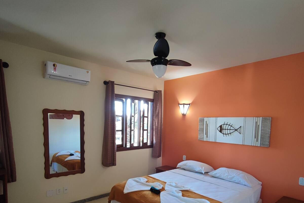 Quarto de hotel com cama, ventoinha de teto, aparelho de ar condicionado, espelho, janela com cortinas e luminária na parede.