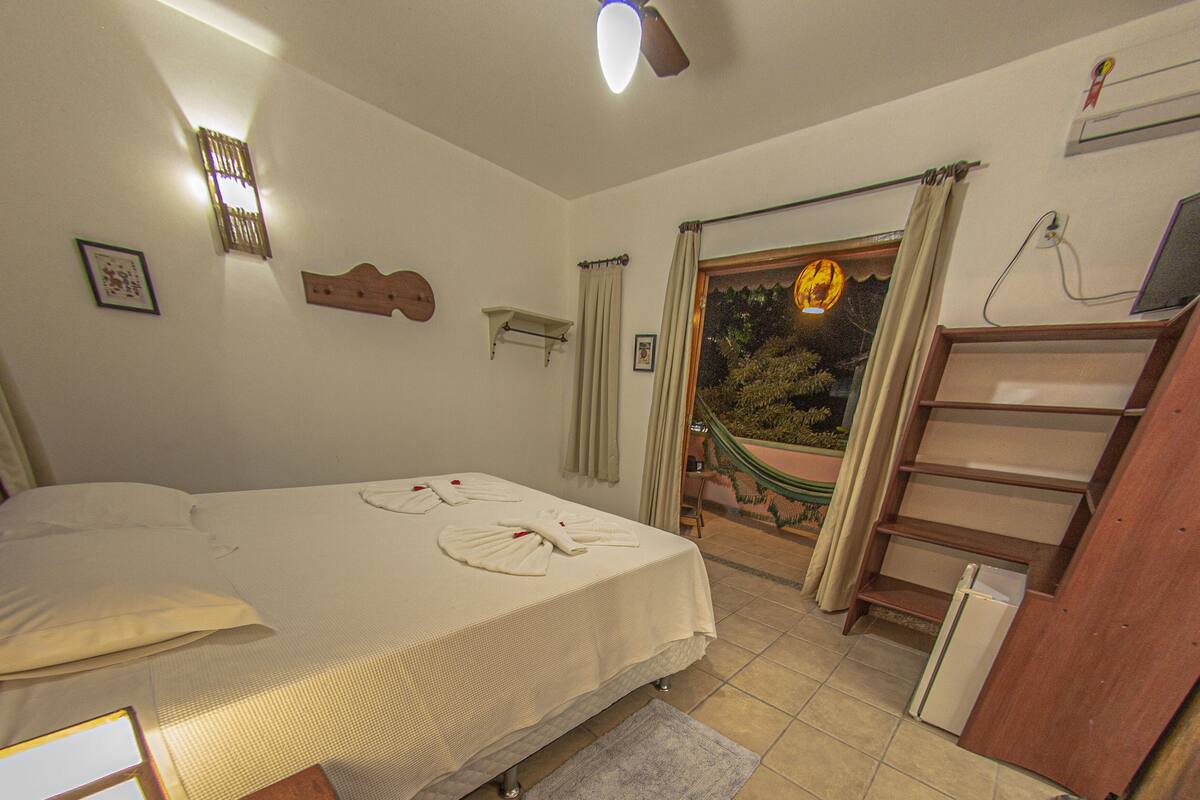 Quarto com cama, uma mesinha, escrivaninha, cadeira, janela com cortinas e vista para o exterior.