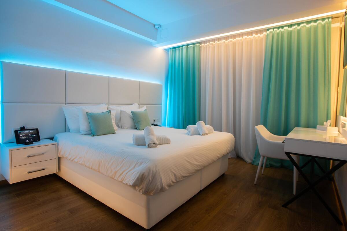 Quarto de hotel moderno com uma cama bem arrumada, mesa de cabeceira com relógio, escrivaninha com cadeira e uma janela grande com cortinas.