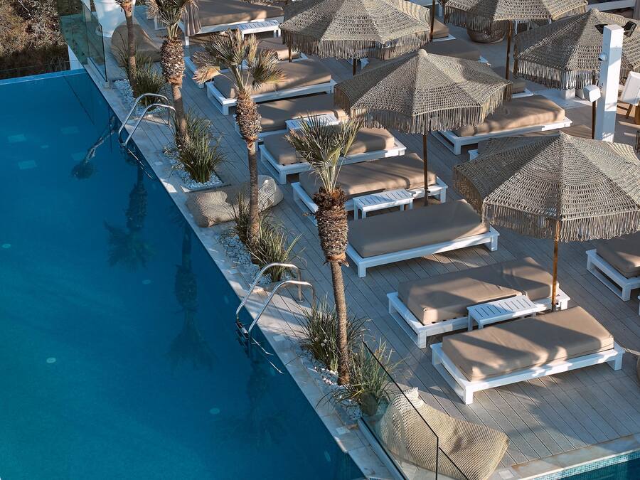 Uma área junto à piscina com espreguiçadeiras, guarda-sóis e palmeiras.