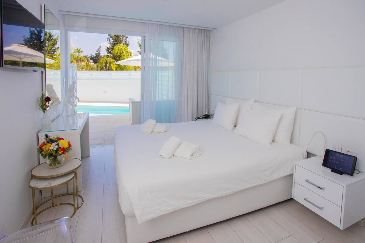 Quarto de hotel moderno com uma cama grande, uma mesinha de cabeceira com relógio digital, uma pequena mesa redonda com um vaso de flores e vista para uma piscina e palmeiras pela janela.