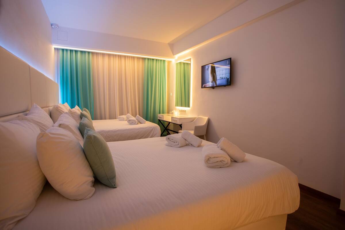 Quarto de hotel com duas camas, uma escrivaninha, uma cadeira, uma televisão e uma janela com cortinas.