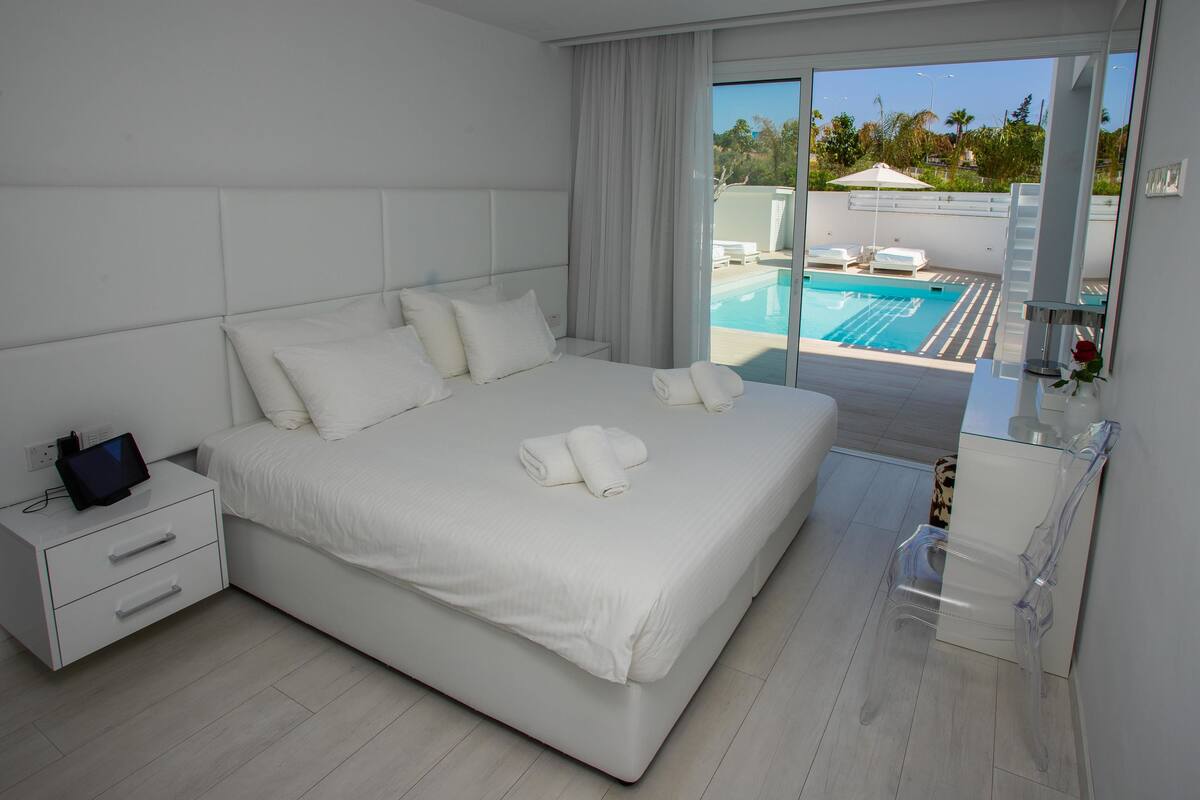Quarto de hotel moderno com uma cama grande, uma mesinha de cabeceira com televisão e vista para uma piscina.