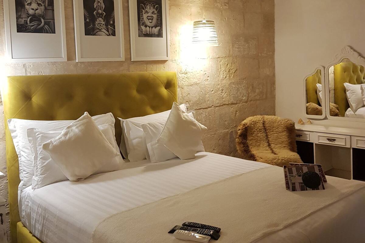 Uma cama com roupa de cama branca, cabeceira amarela e mesa de cabeceira com abajur. Há quadros nas paredes e um espelho sobre a cama.