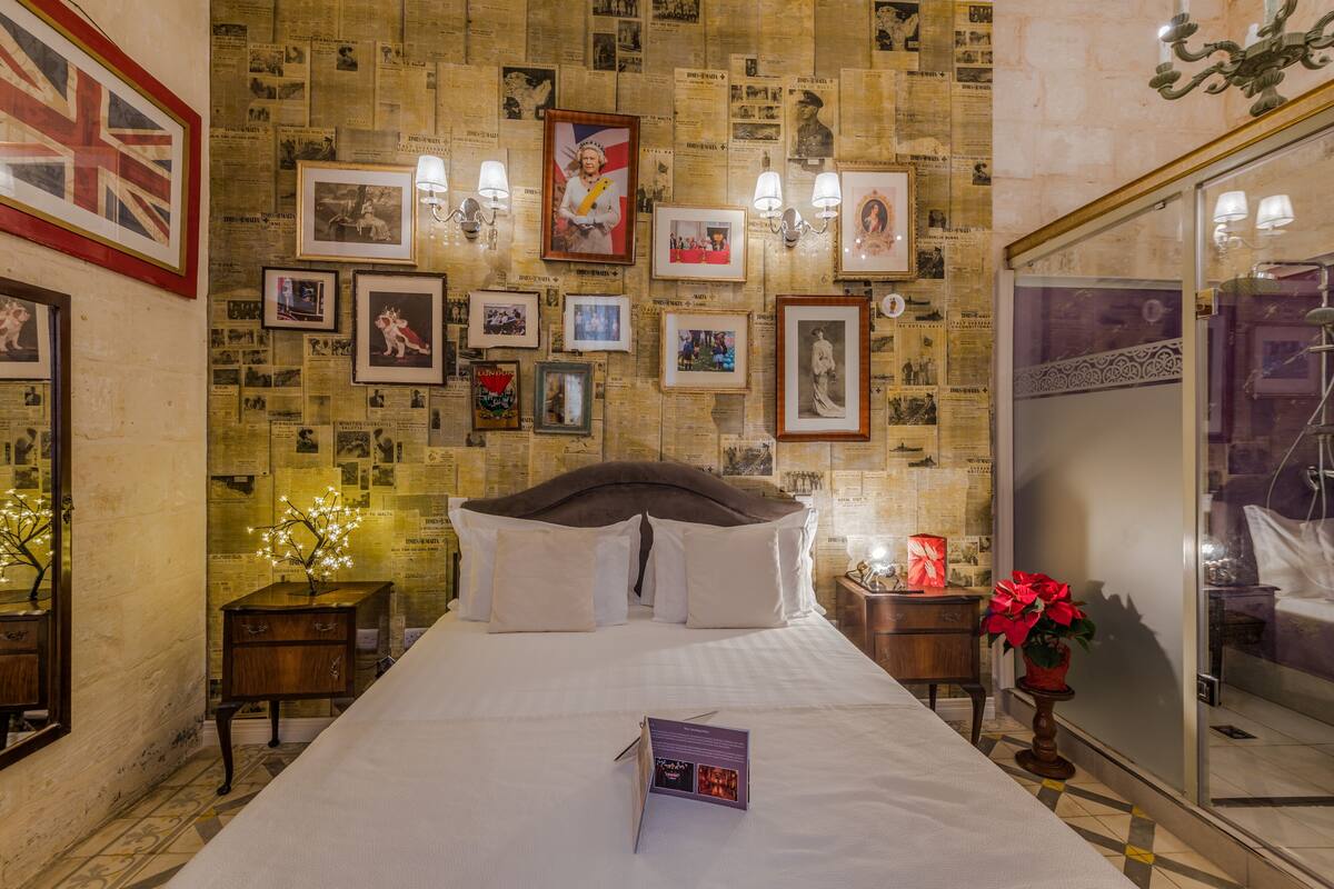 Quarto de hotel com uma cama grande, um espelho, uma mesa de cabeceira e uma parede decorada com quadros e jornais.