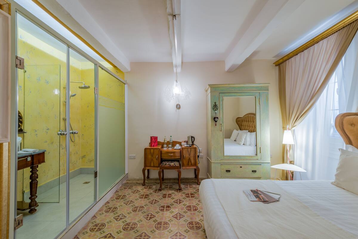 Quarto de hotel com cama, escrivaninha com espelho, chuveiro e piso estampado.