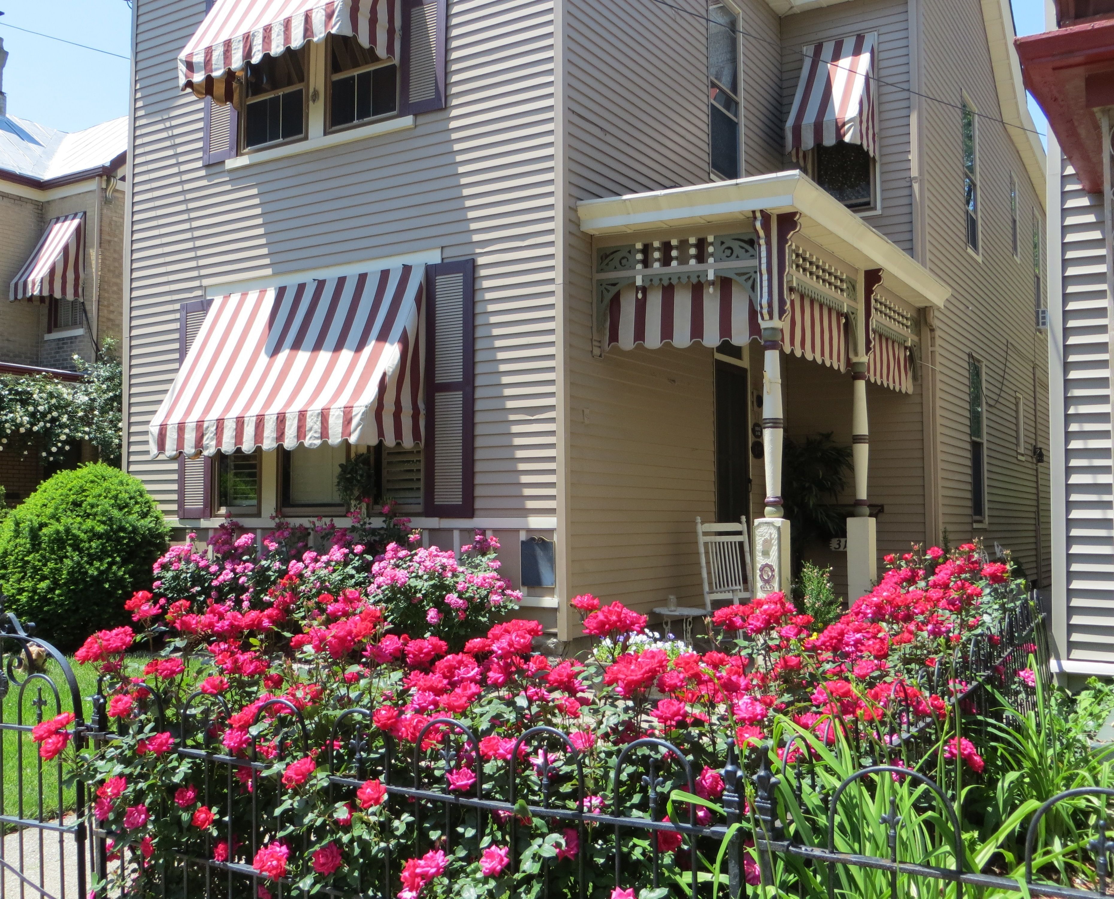 Uma casa de dois andares com varanda, toldos listrados e um jardim com flores cor-de-rosa.