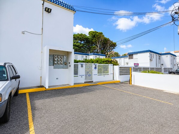 Uma área de estacionamento com um edifício branco, entrada com portão e um veículo.