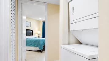 Lavanderia com máquina de lavar e secadora, uma cama no quarto vizinho e uma porta que dá para outra área.