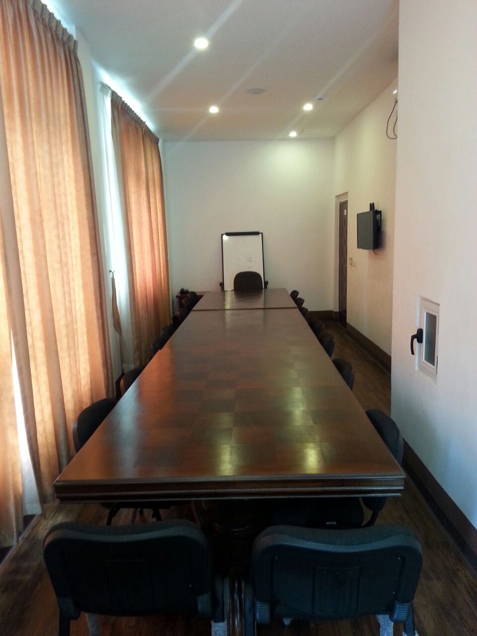 Uma sala de conferências com uma mesa comprida, cadeiras, um quadro branco e uma televisão na parede.