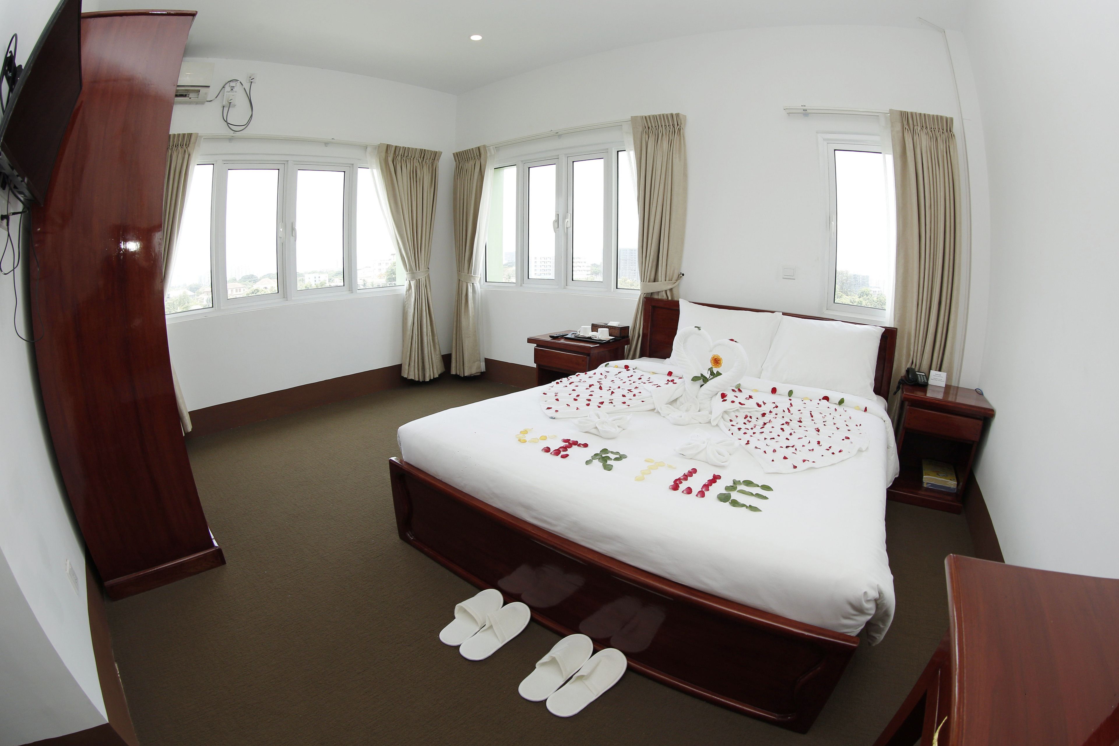 Quarto de hotel com uma cama grande, duas mesas de cabeceira, um guarda-roupa e duas janelas com cortinas.