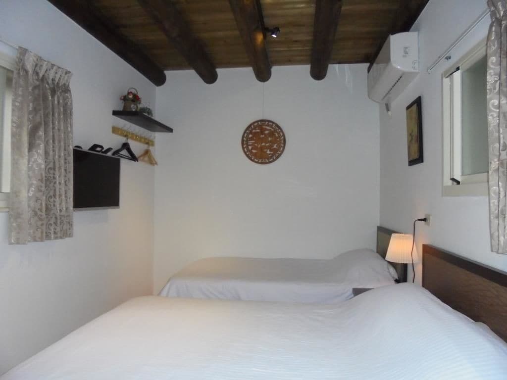 Quarto com duas camas, teto de madeira, aquecedor fixado na parede, janela com cortinas e relógio de parede.