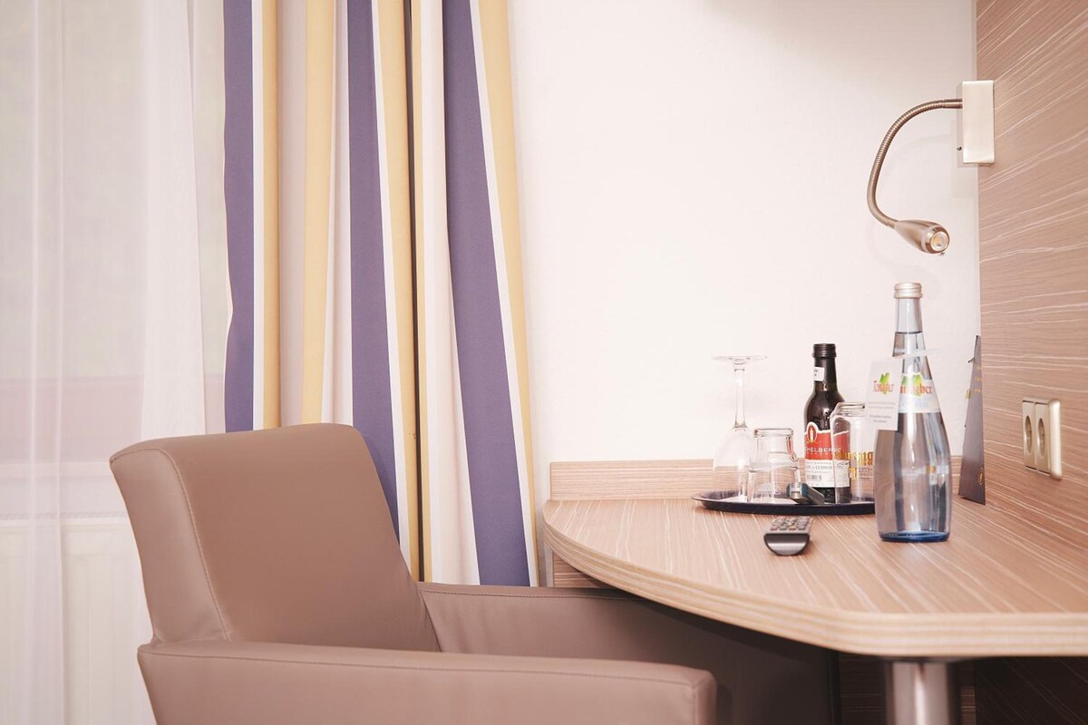 Quarto de hotel com mesa de madeira curvada, uma poltrona de couro, minibar e prateleira com diversos itens.