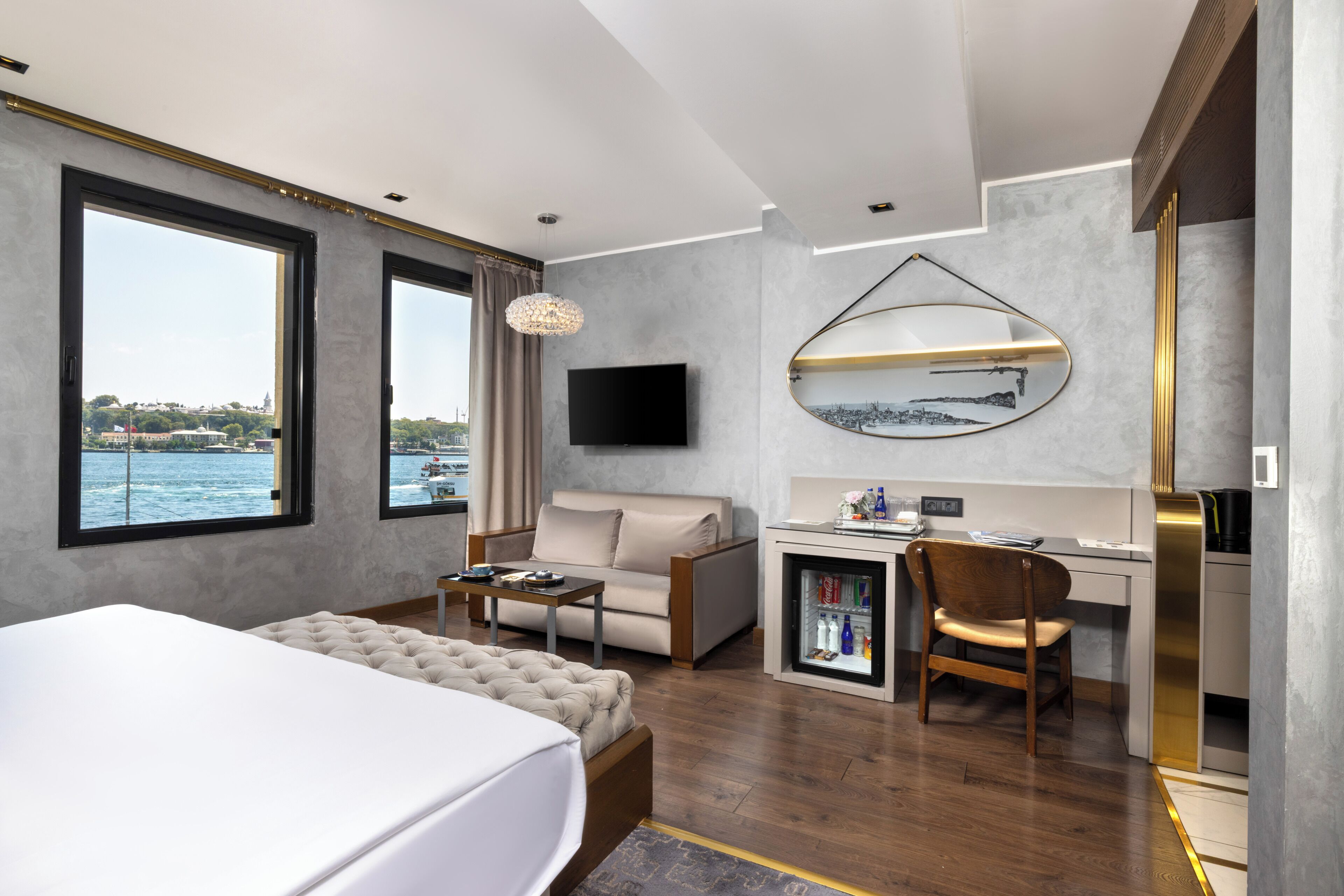 Quarto de hotel moderno com uma cama grande, área de estar, escrivaninha, televisão e vista para o mar.