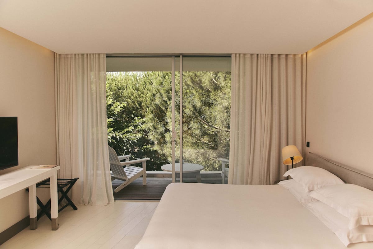 Quarto de hotel com cama, escrivaninha, cadeira, mesa e vista para árvores através de uma grande janela.