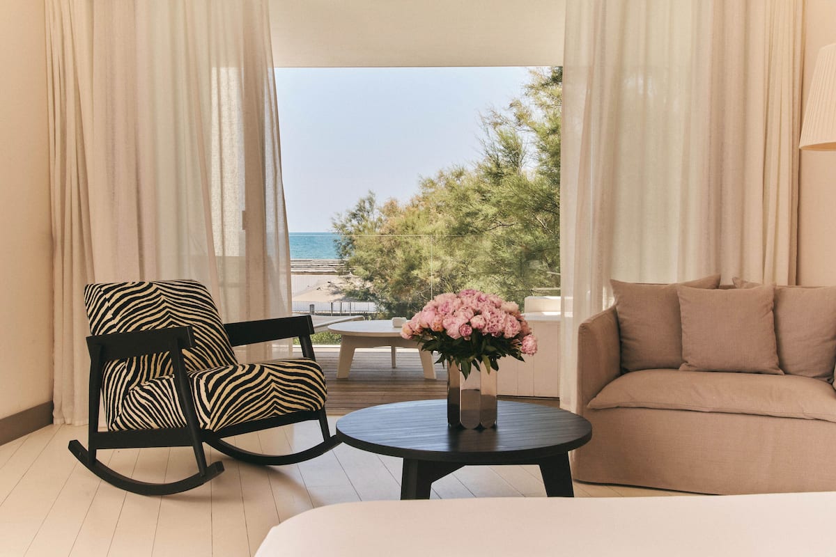 Um quarto com uma poltrona estampada em zebra, um sofá bege, uma pequena mesa redonda com um vaso de flores e vista para o mar através de grandes janelas.