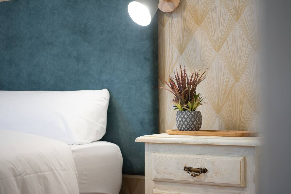 Uma cama bem arrumada com roupa de cama branca, uma pequena mesa de cabeceira com uma planta e uma parede com padrão geométrico.