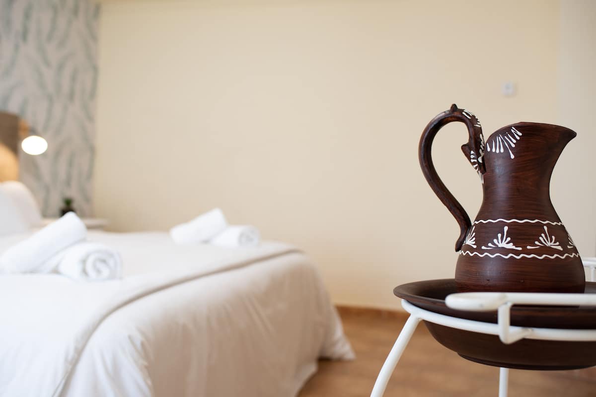 Quarto de hotel com cama, toalhas dobradas com cuidado e um jarro de cerâmica decorativo sobre um suporte branco.