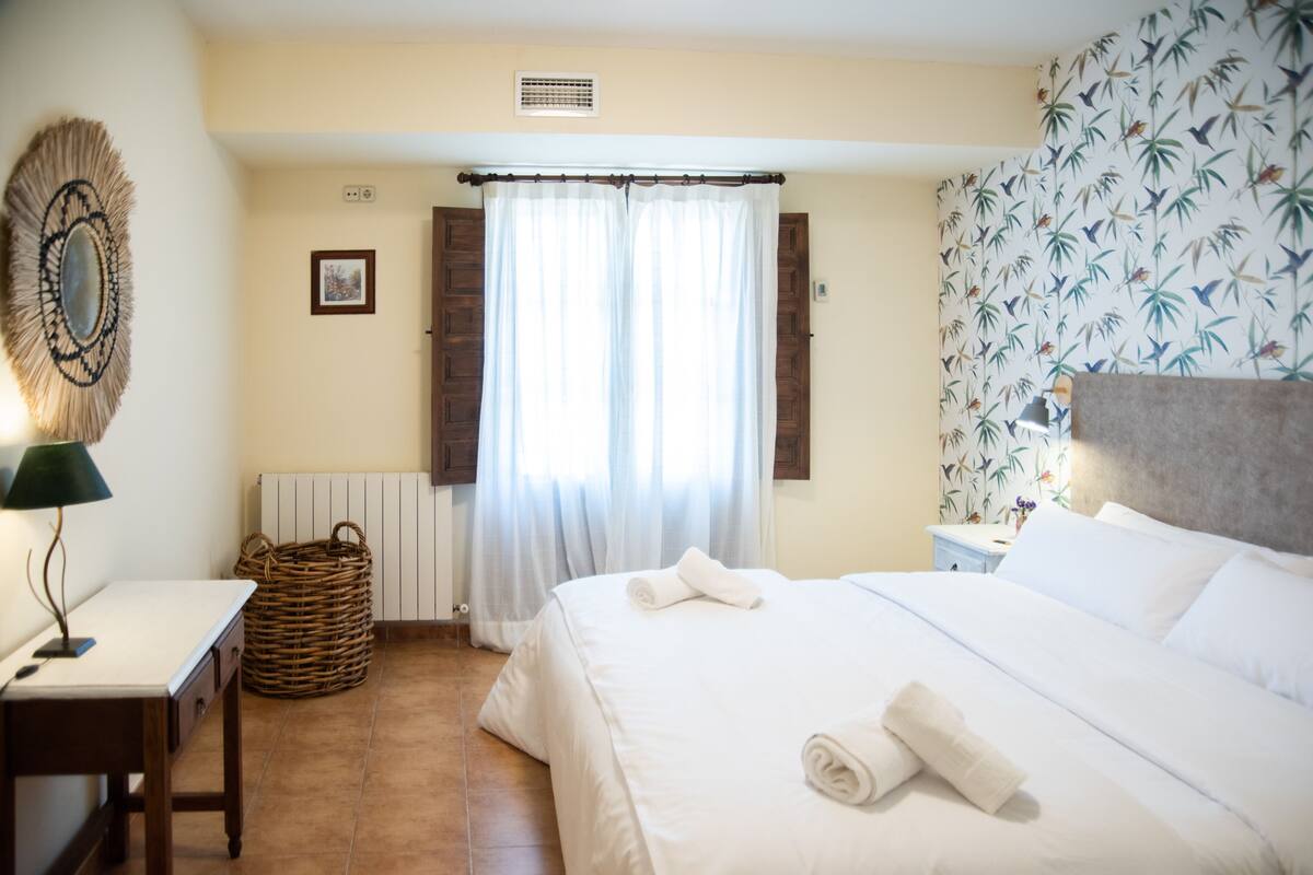 Um quarto com cama, escrivaninha, luminária, cesta de vime e janela com cortinas.