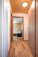 Um corredor estreito com piso de madeira e teto com um claraboia circular. Uma porta à direita dá acesso a um quarto com cama e uma mesinha.
