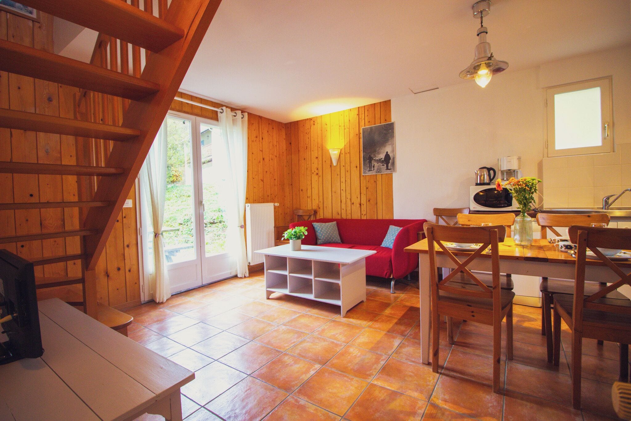 Um quarto acolhedor com escada de madeira, um sofá vermelho, uma mesa de centro branca e um espaço de refeições com cadeiras e mesa de madeira.
