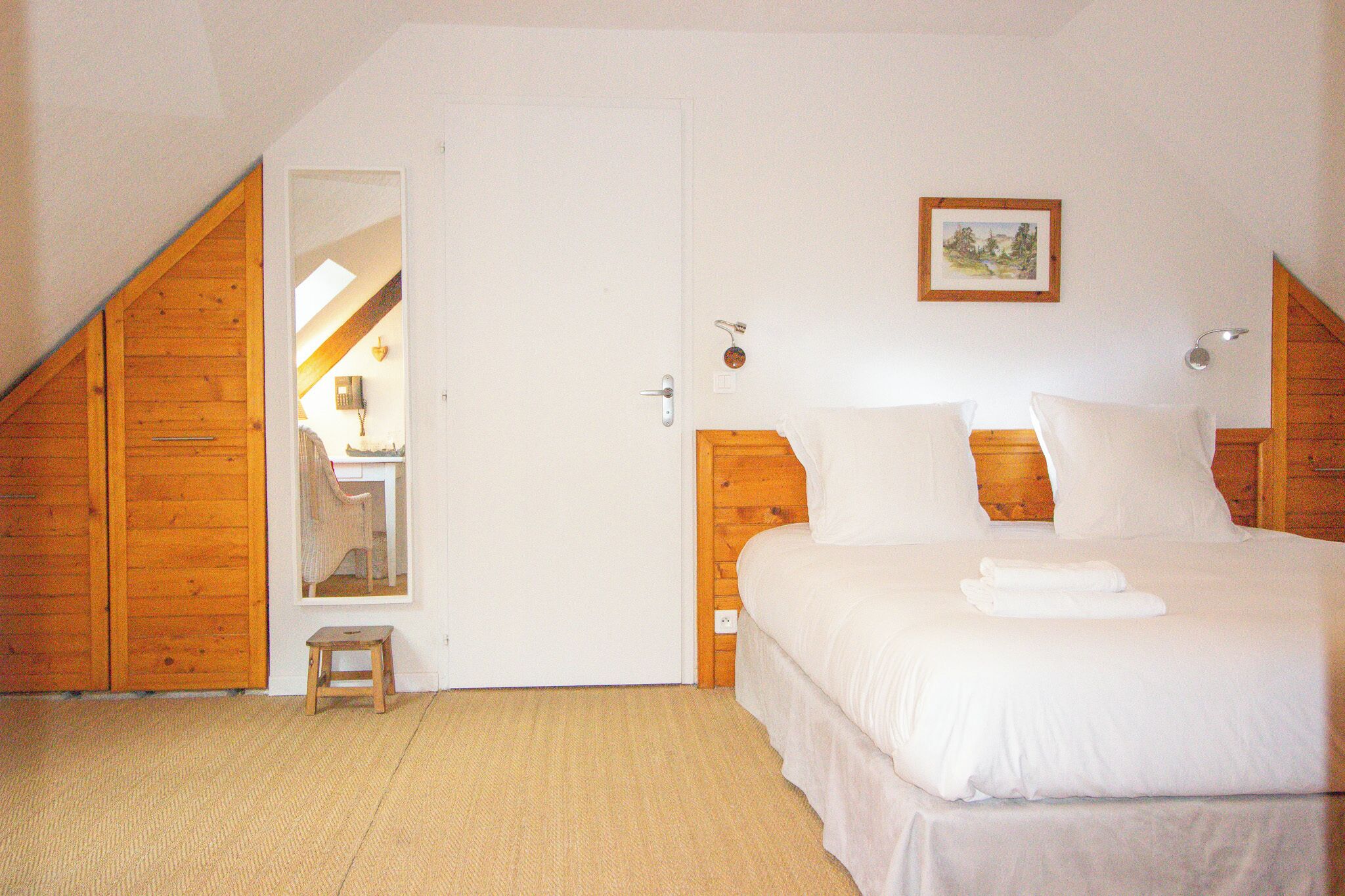 Quarto com cama de madeira, um banquinho pequeno, porta branca e teto inclinado.