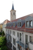 Um edifício de vários andares com telhado de telhas vermelhas, uma torre imponente e uma extensão moderna com varandas.
