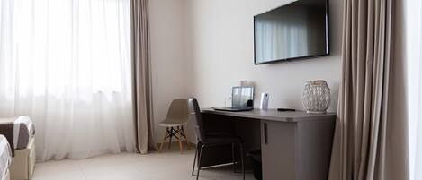 Quarto de hotel com cama, escrivaninha, cadeira, televisão e janela com cortinas.