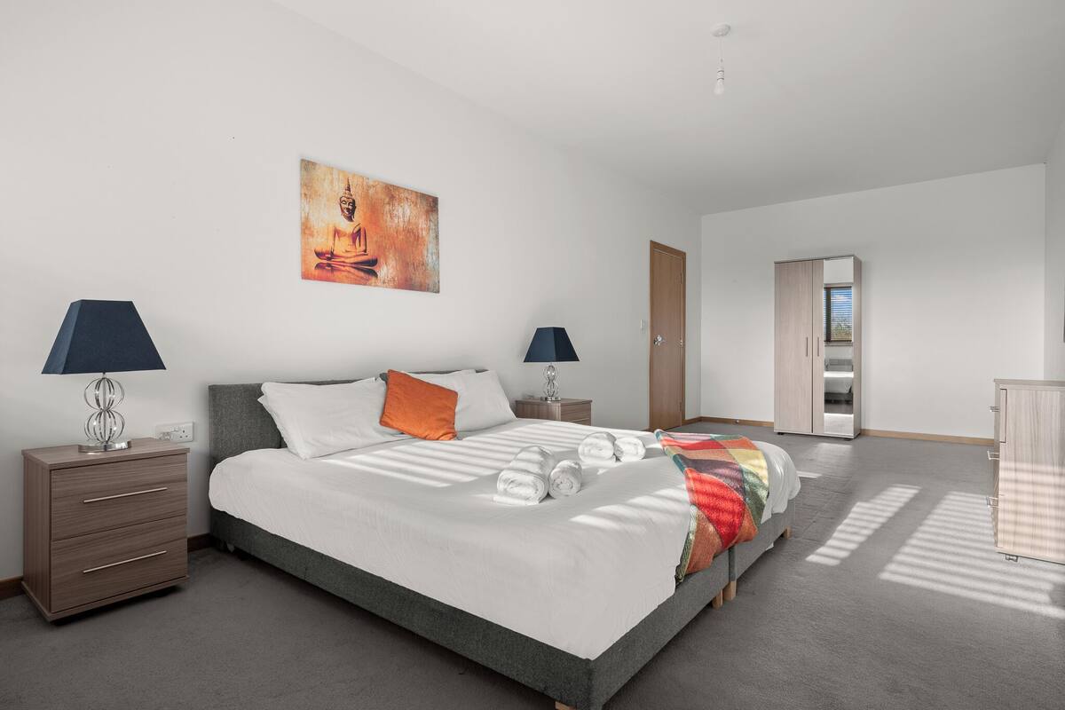 Quarto de hotel moderno com uma cama grande, duas mesas de cabeceira com abajures, um guarda-roupa e um quadro na parede.