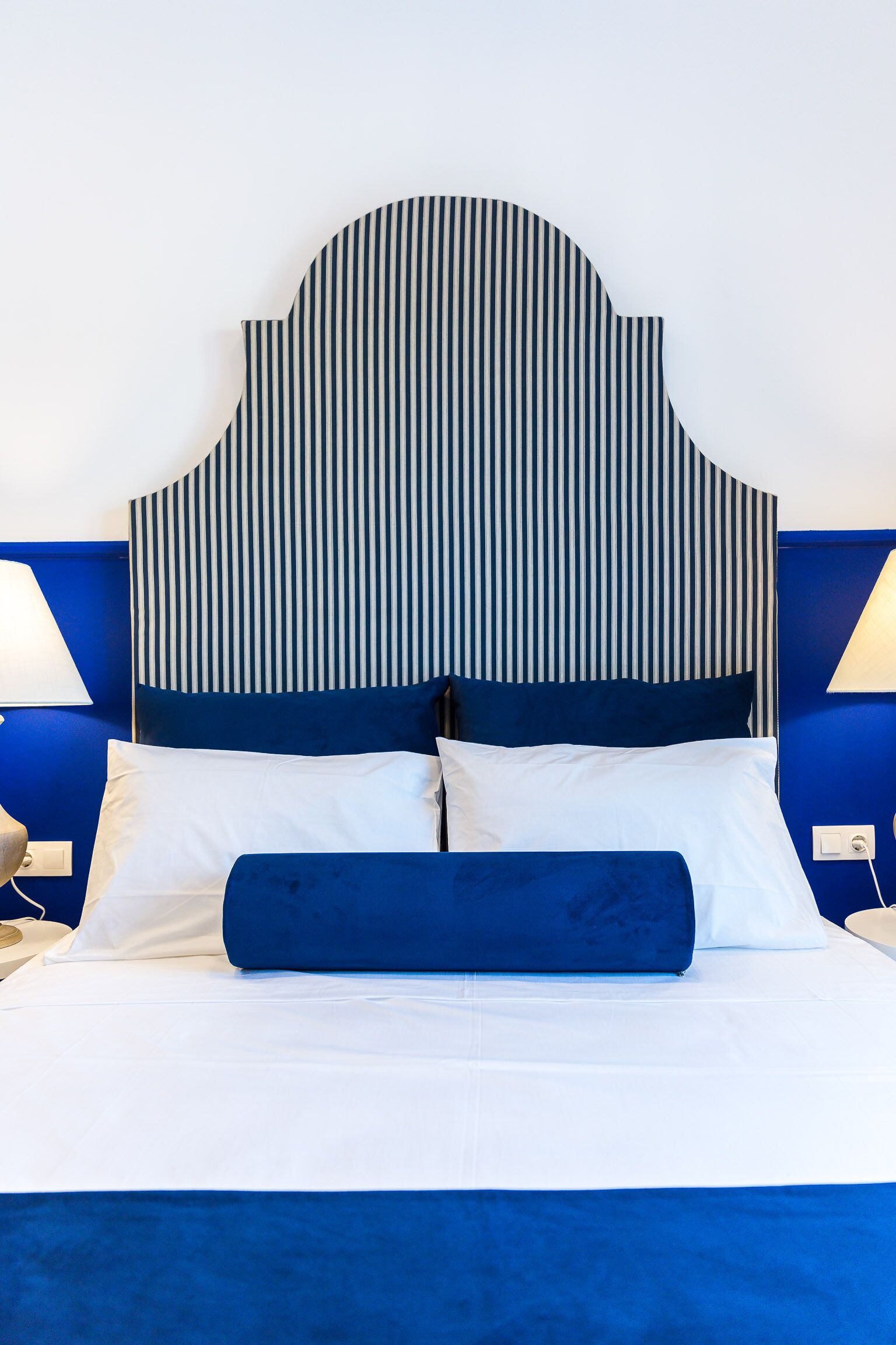Uma cama com cabeceira listrada, roupa de cama branca e azul, e duas luminárias de apoio.