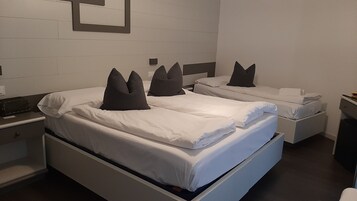 Quarto de hotel com duas camas, cada uma com lençóis brancos e almofadas escuras. Há uma pequena mesa de cabeceira com uma luminária em cada cama. As paredes são brancas, com uma parede de destaque cinza atrás das camas.