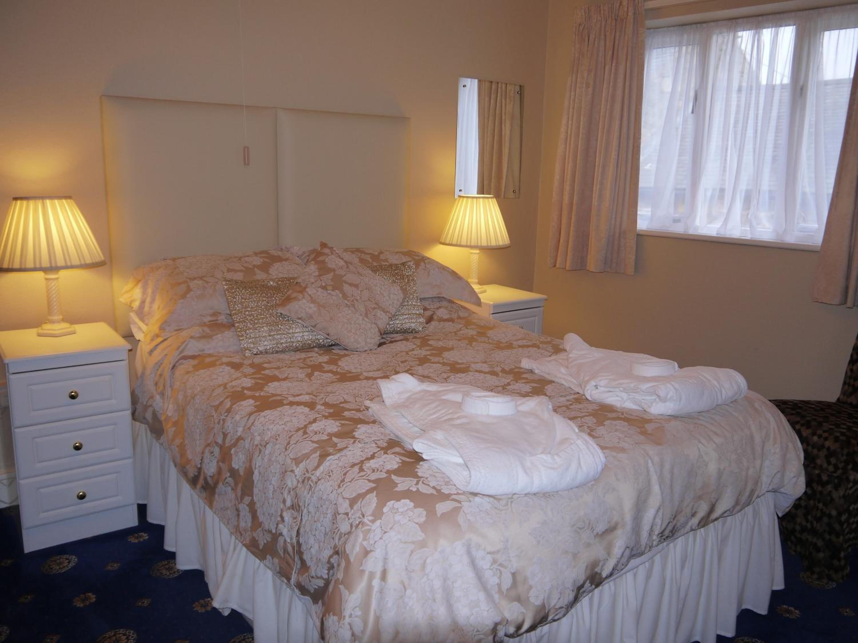 Uma cama bem arrumada com colcha floral, duas luminárias de cabeceira, uma mesa de cabeceira com gaveta e uma janela com cortinas.