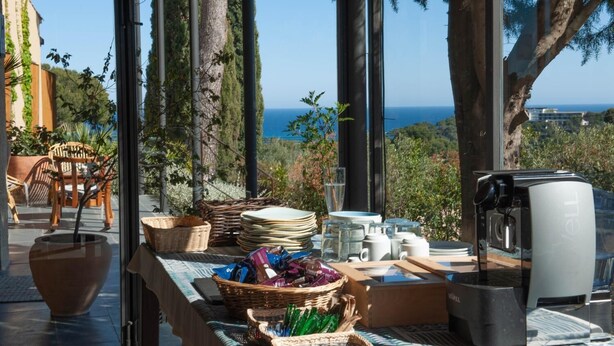 Disposição para o café da manhã com pratos, xícaras e chaleira sobre uma mesa, com vista para o mar e árvores.
