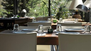 Uma área de refeições com mesas preparadas para dois, equipada com copos, talheres e uma garrafa. Grandes janelas oferecem vista para uma paisagem arborizada.