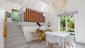 Um interior moderno com uma escada, uma área de jantar com mesa de vidro e cadeiras brancas, e vista para o verde lá fora.