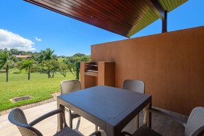 Um pátio externo com mesa e cadeiras, uma pérgola de madeira e vista para uma paisagem verde com árvores e um edifício ao longe.