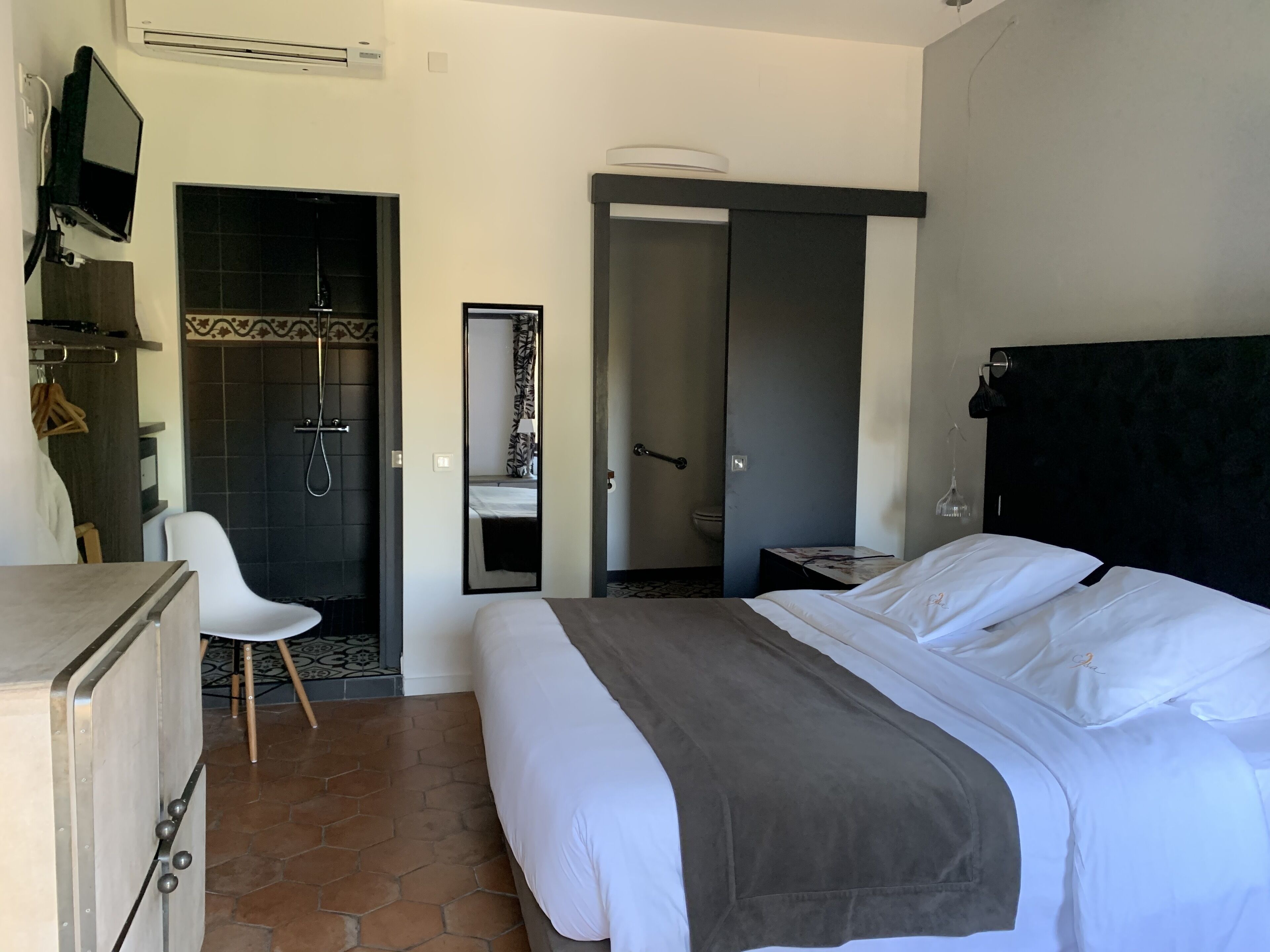 Quarto de hotel com cama, escrivaninha, cadeira, espelho e área de chuveiro.