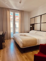 Quarto de hotel com cama, escrivaninha, cadeira e varanda com cortinas.