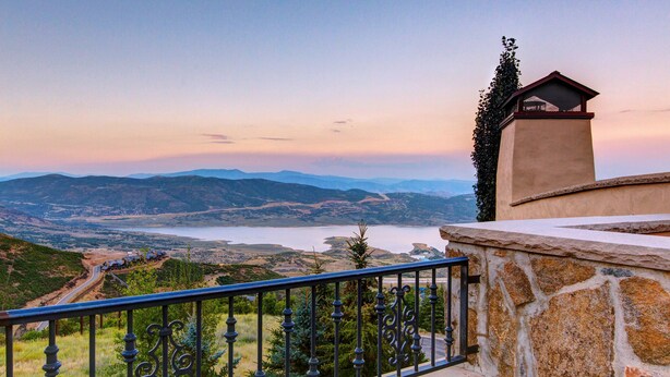 Varanda com muro de pedra e grade de metal, com vista para um vale, um lago e montanhas ao longe.