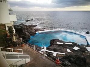 Uma piscina com água azul cristalina, rodeada por terreno rochoso e com vista para o oceano.